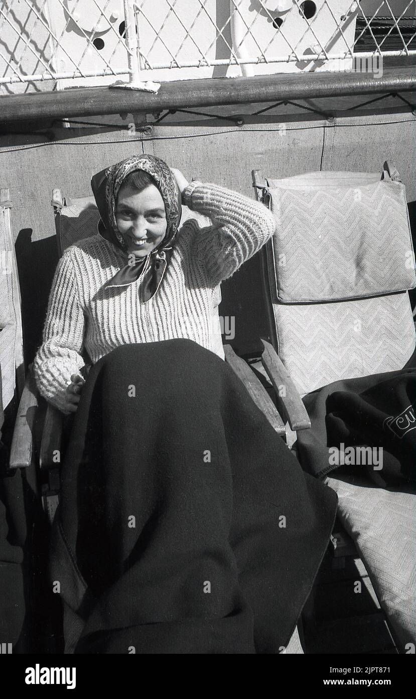 1950s, historical, a female ship passenger wearing sweater and head scarf sitting outside on a ...