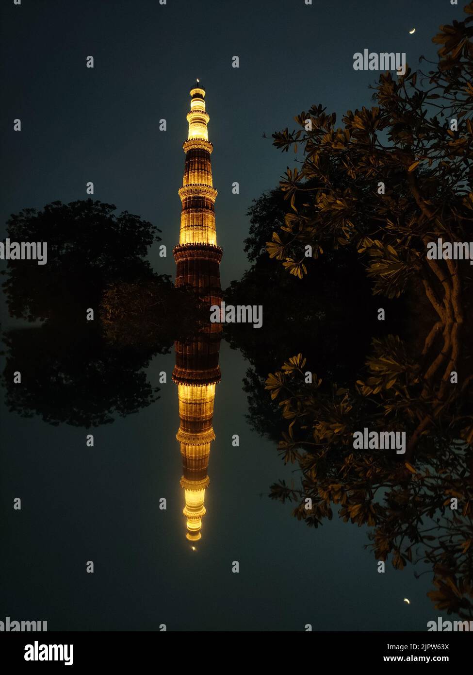 A vertical shot of the Qutb Minar complex and its reflection in the water at night in Delhi ...