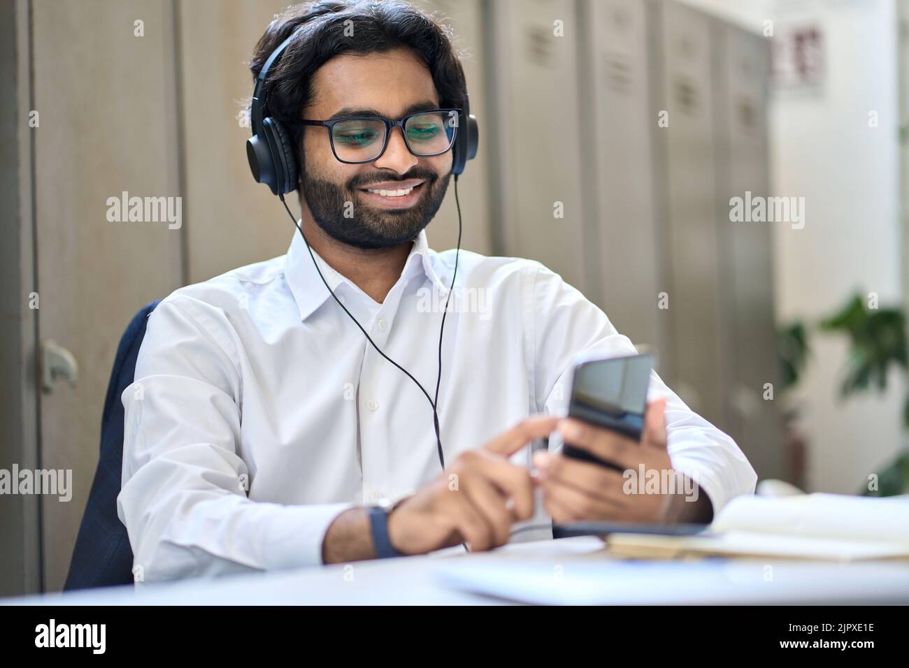 Smiling indian business man wearing headphones using smartphone at work ...