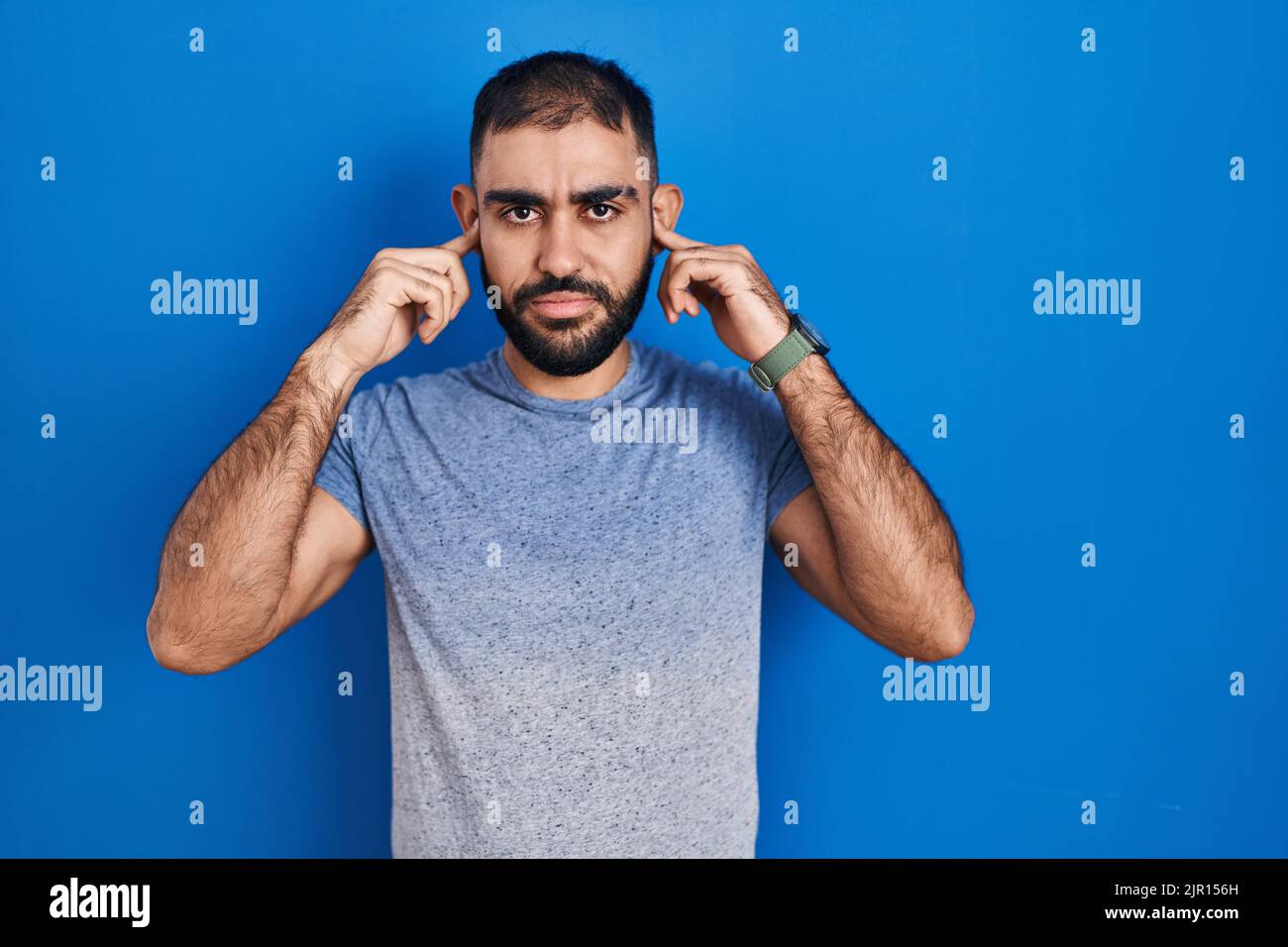 Middle east man with beard standing over blue background covering ears ...