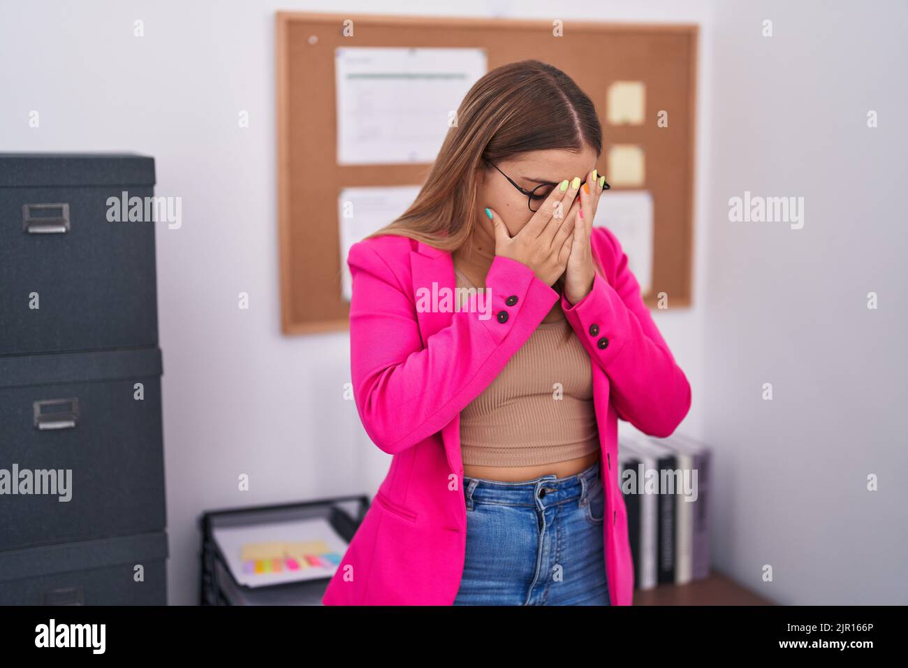 Young blonde woman standing at the office with sad expression covering ...