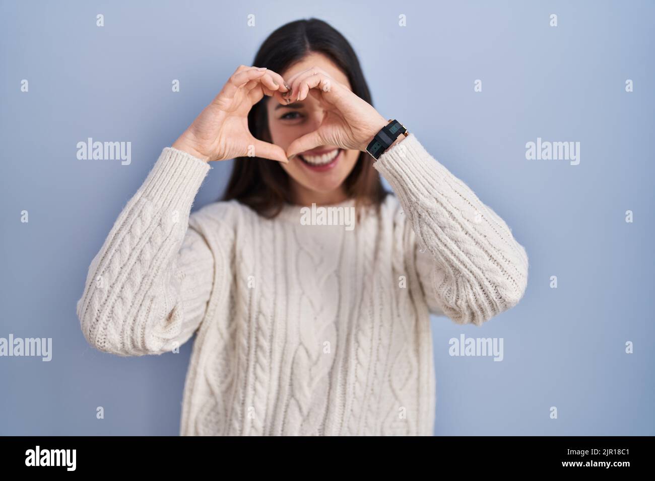 Young brunette woman standing over blue background doing heart shape ...