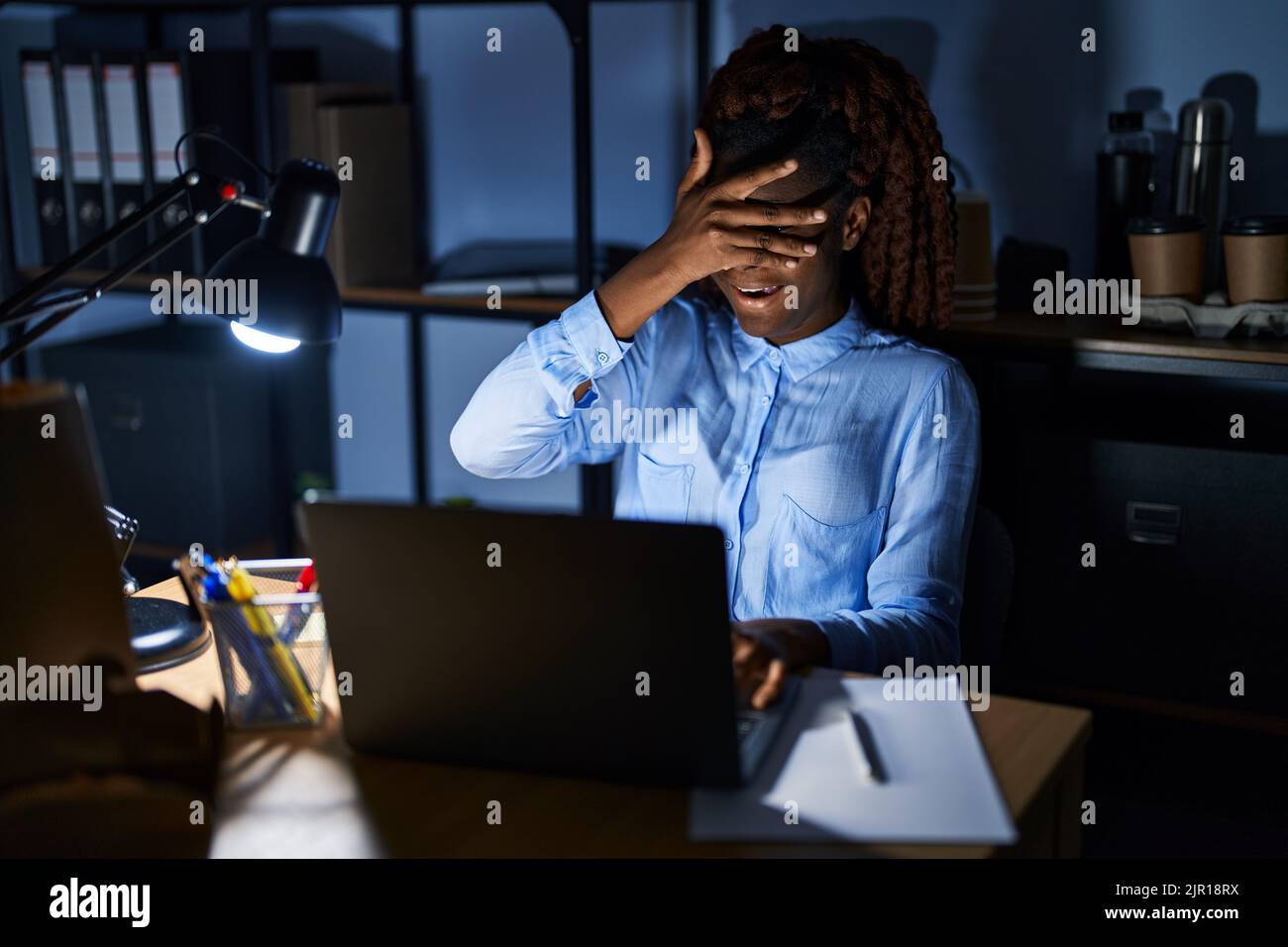 African woman working at the office at night peeking in shock covering ...