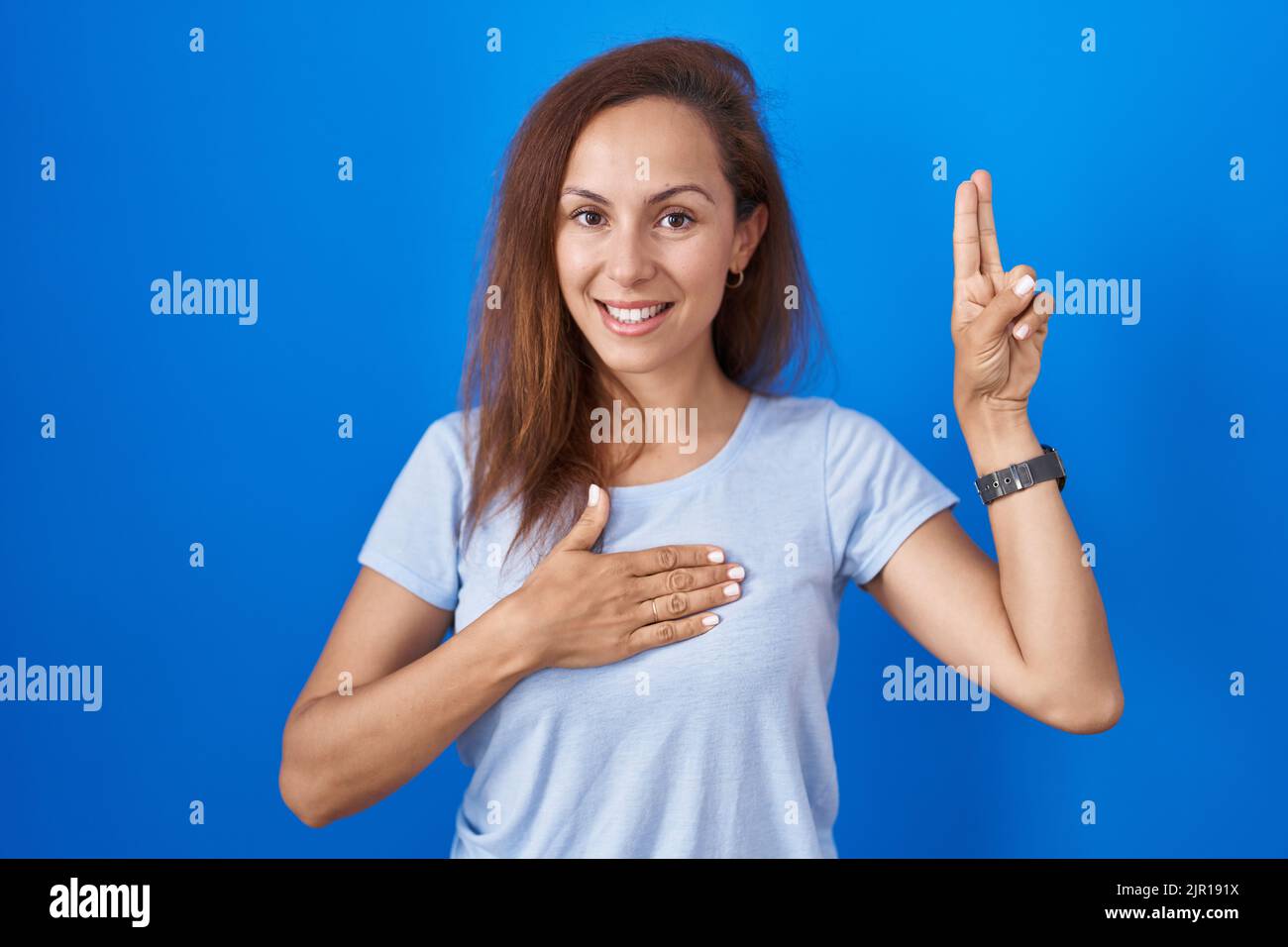 Brunette woman standing over blue background smiling swearing with hand ...