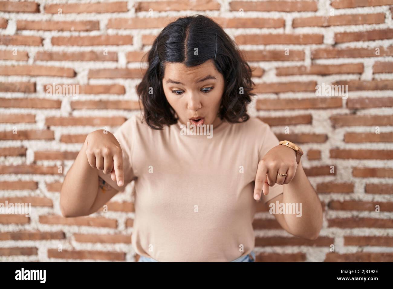 Young hispanic woman standing over bricks wall pointing down with fingers showing advertisement ...