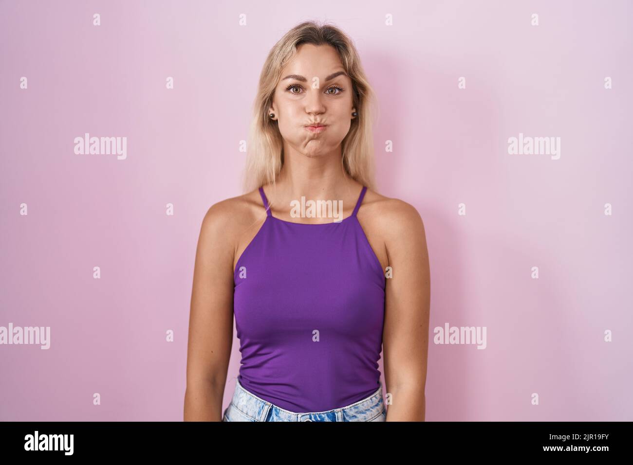 Young blonde woman standing over pink background puffing cheeks with ...