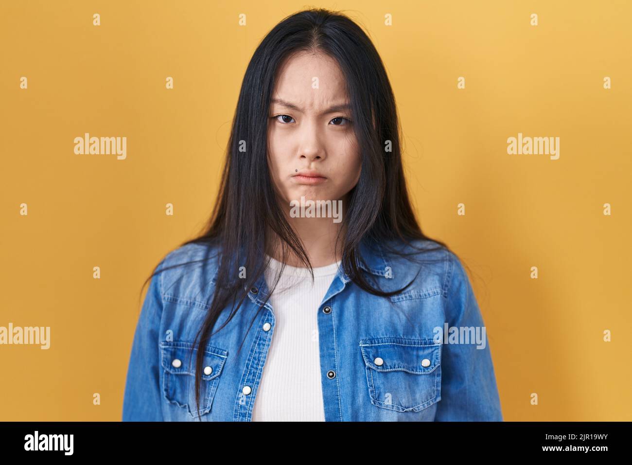Young asian woman standing over yellow background skeptic and nervous ...