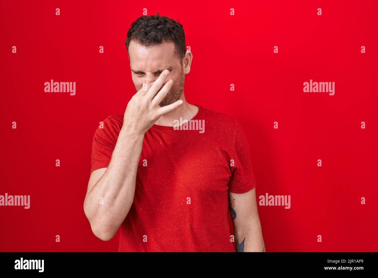 Young hispanic man standing over red background smelling something ...
