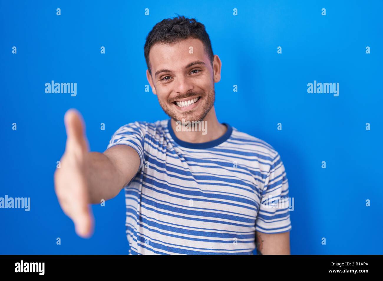 Young hispanic man standing over blue background smiling friendly ...