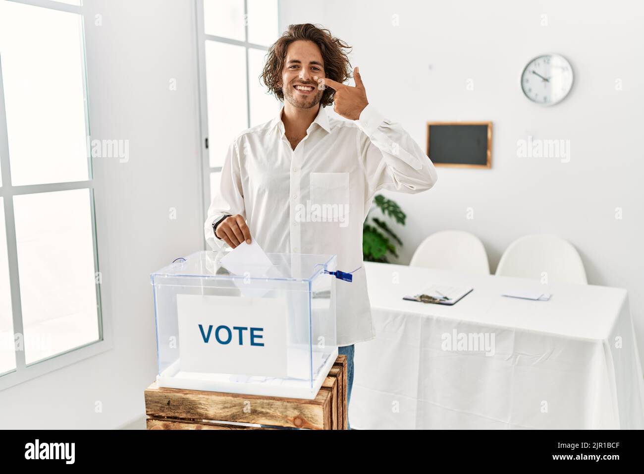 Young hispanic man voting putting envelop in ballot box pointing with ...