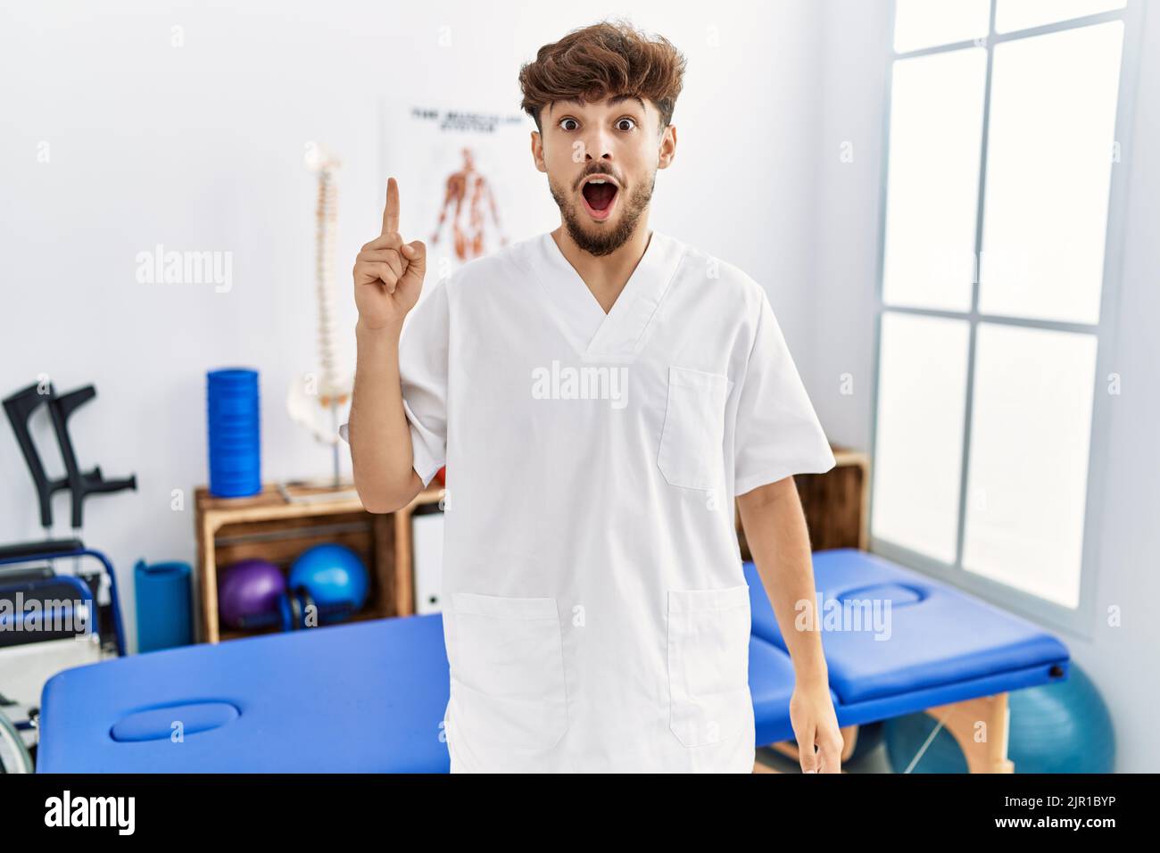 Young arab man working at pain recovery clinic pointing finger up with ...