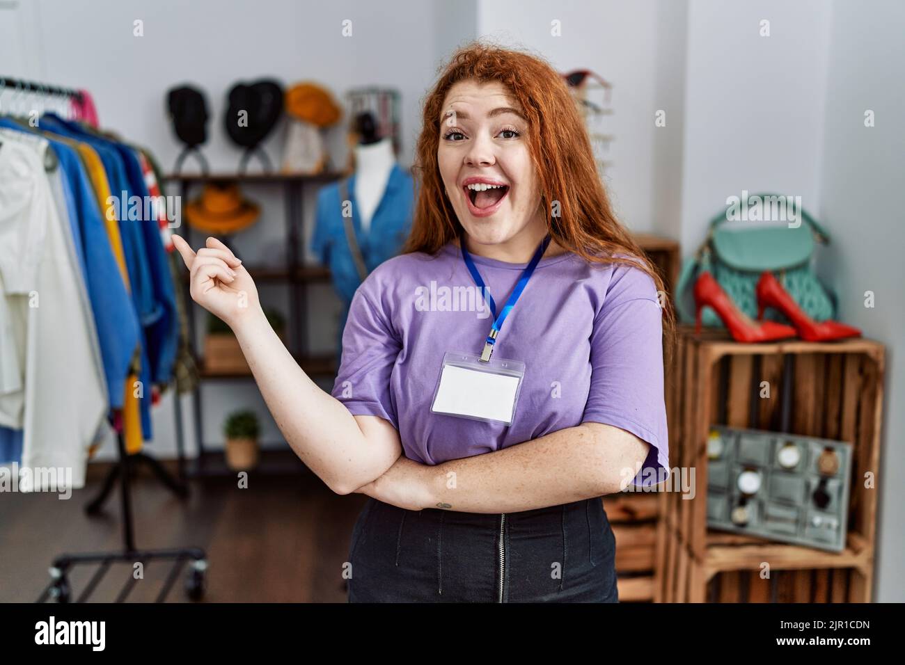 Young redhead woman working as manager at retail boutique with a big smile on face, pointing ...