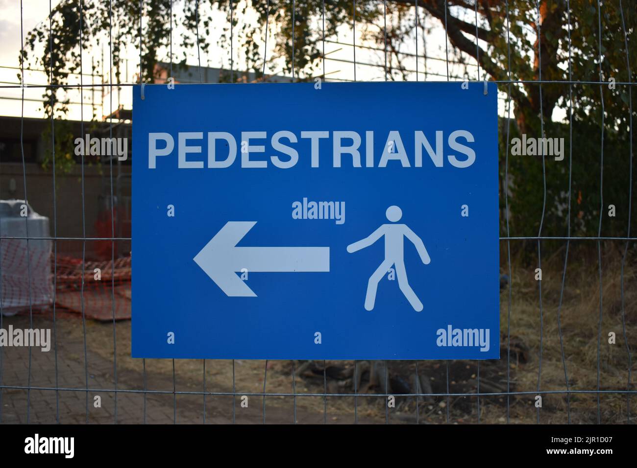 A blue and white sign on a wire fence: "Pedestrians" with a figure and ...