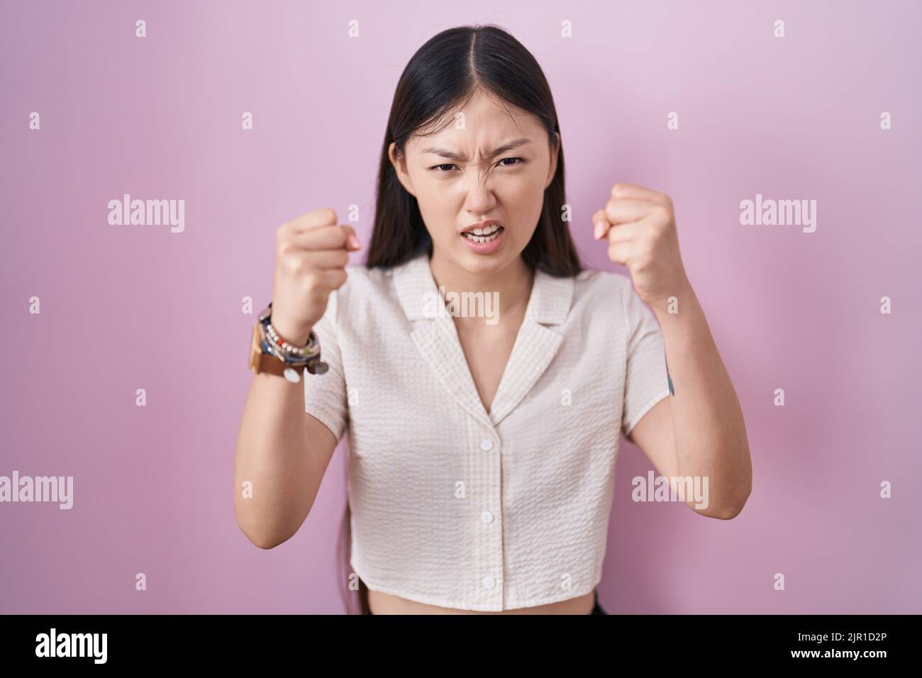 Chinese young woman standing over pink background angry and mad raising ...