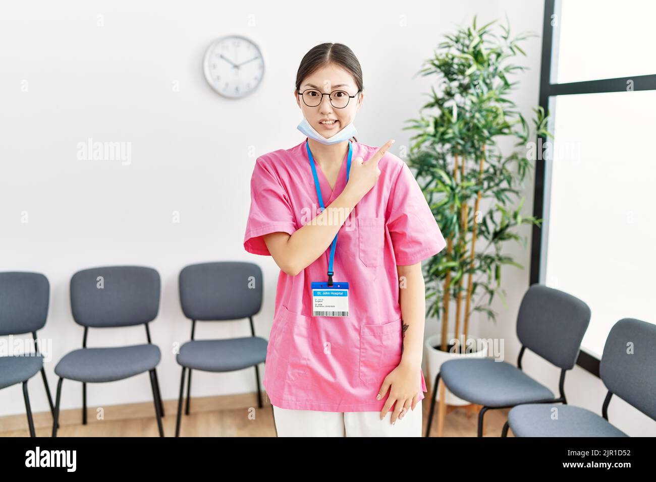 Young asian nurse woman at medical waiting room pointing aside worried ...