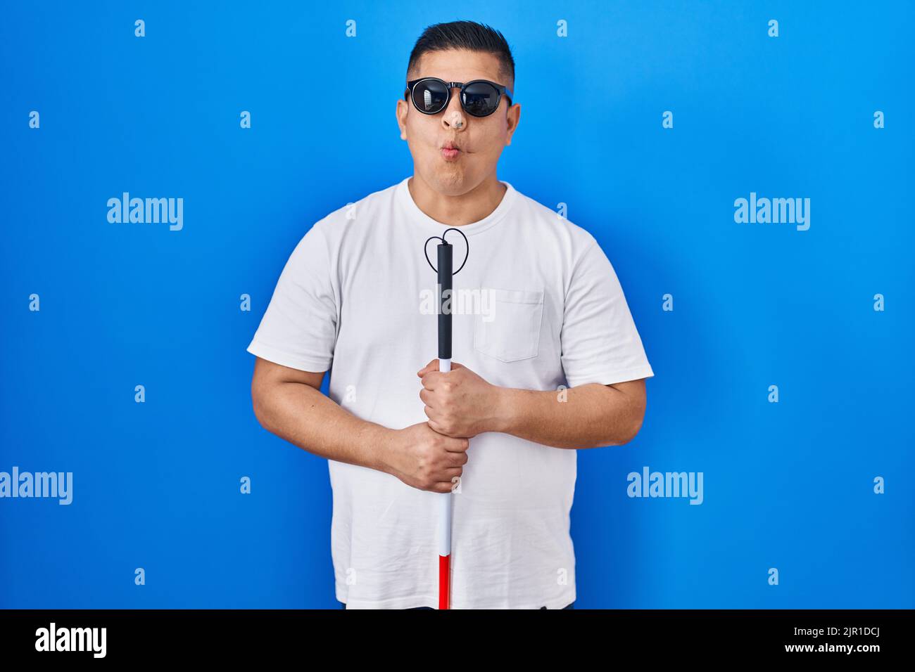 Hispanic young blind man holding cane making fish face with mouth and ...