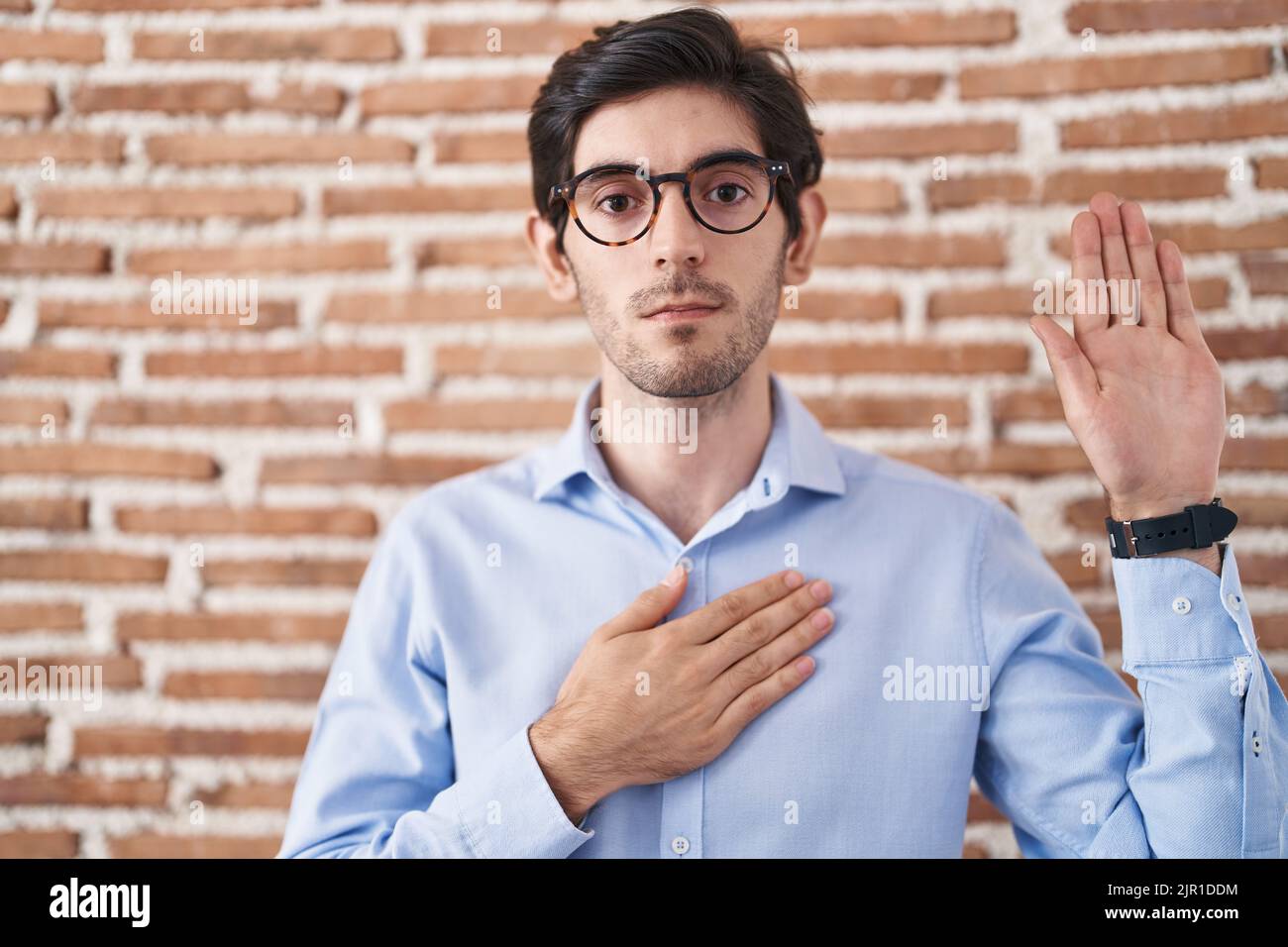 Young hispanic man standing over brick wall background swearing with ...