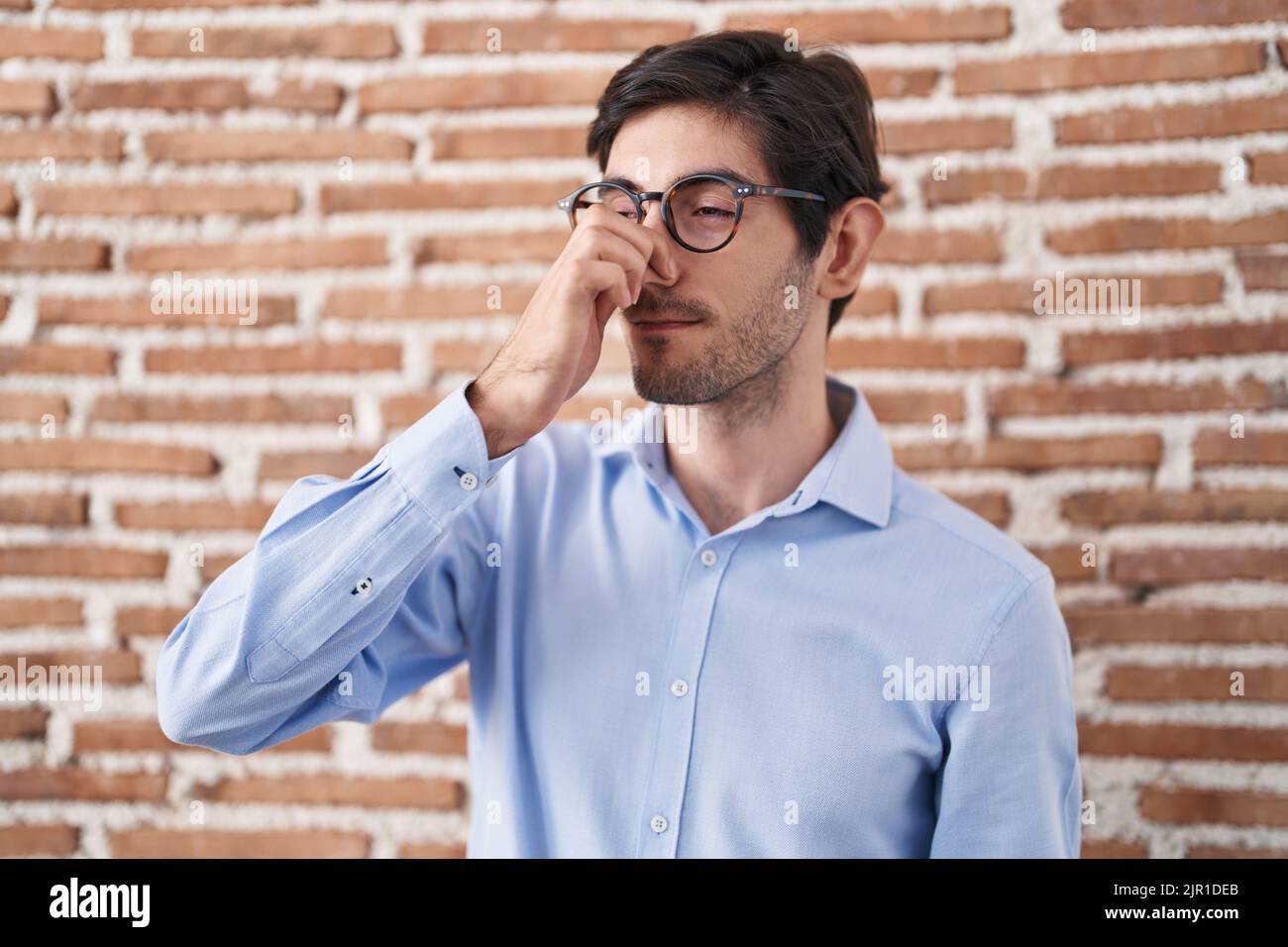 Young hispanic man standing over brick wall background smelling ...