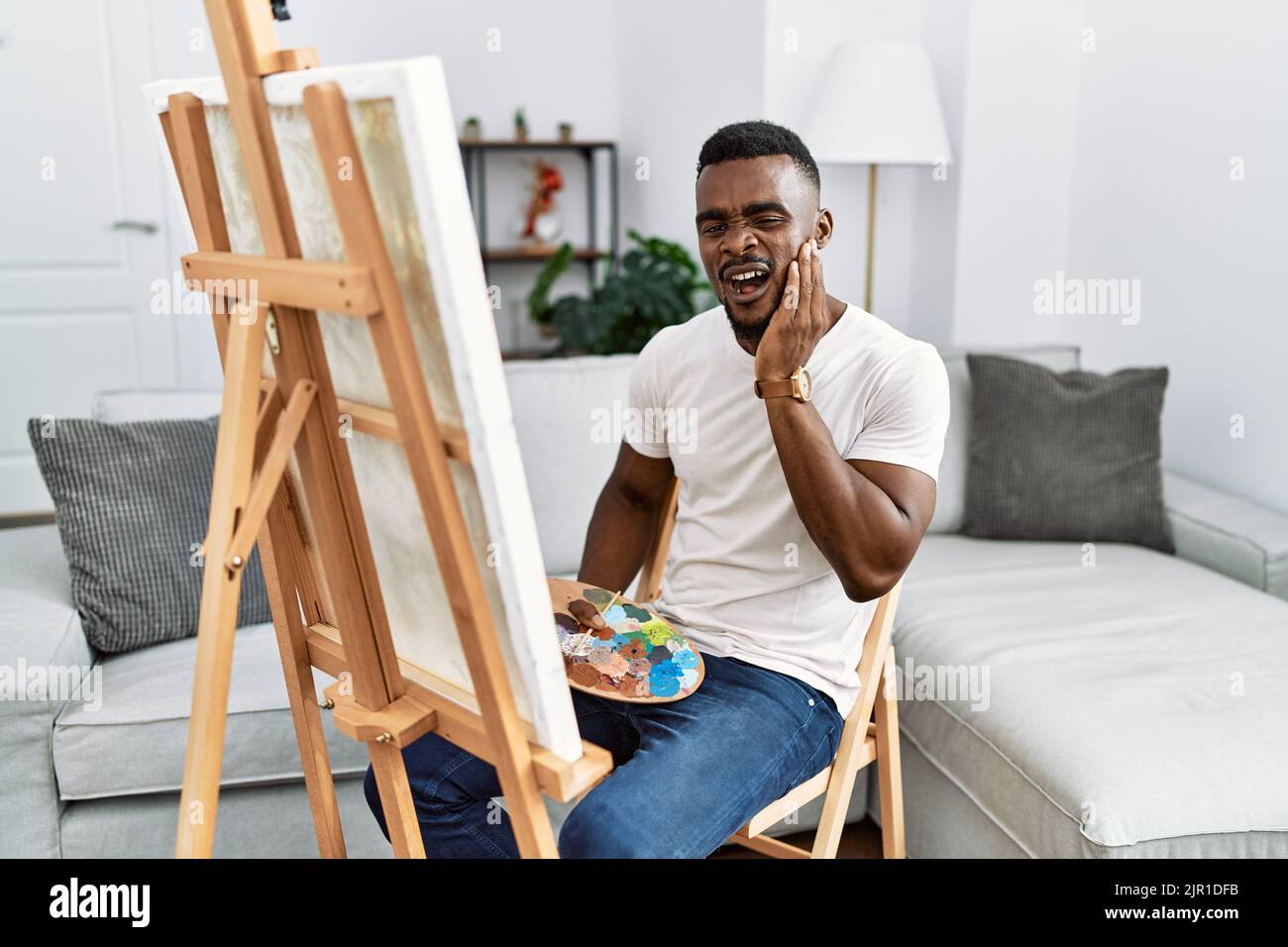 Young african man painting on canvas at home touching mouth with hand ...