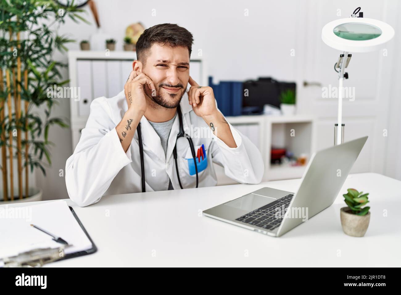 Young doctor working at the clinic using computer laptop covering ears ...