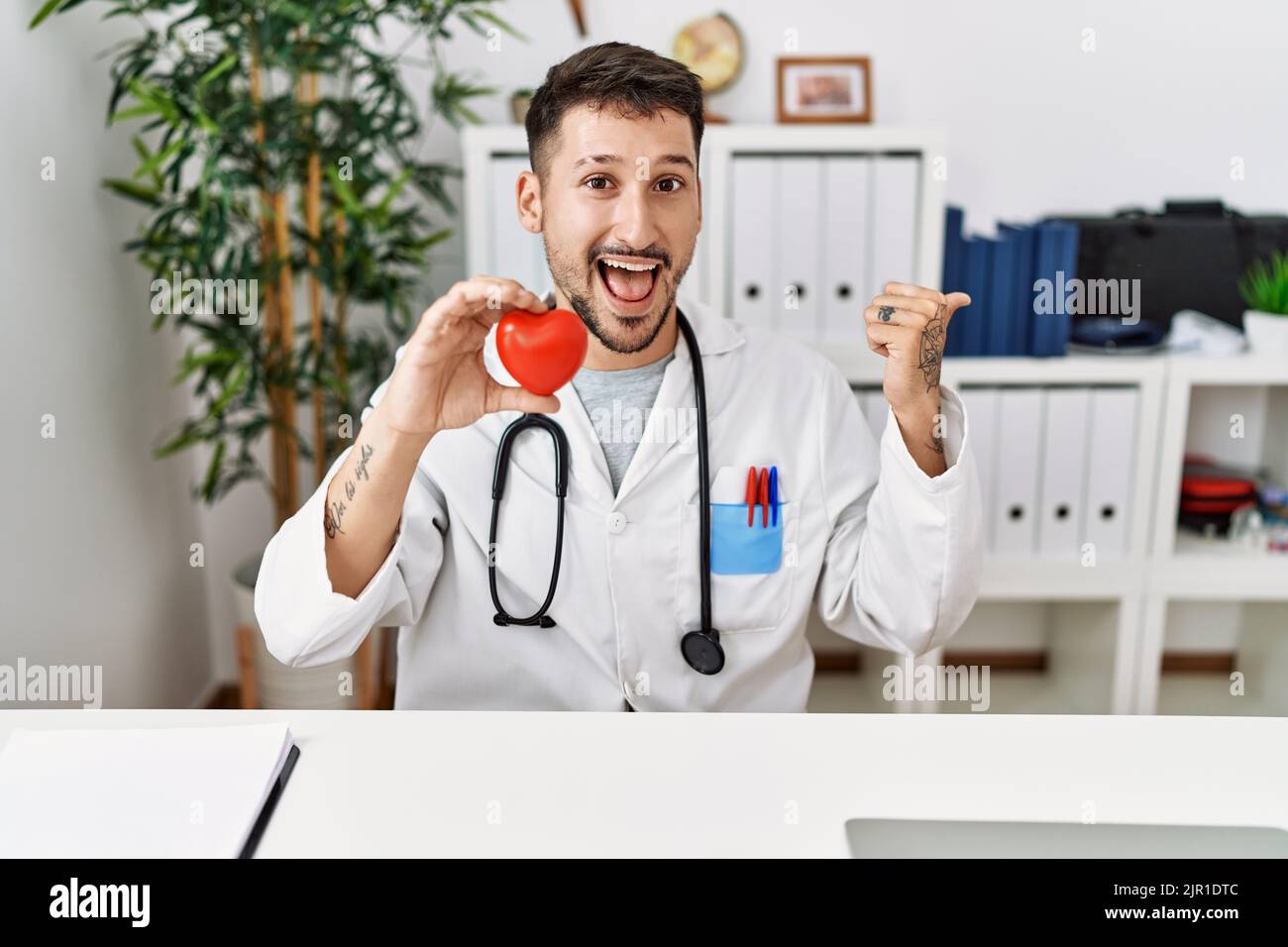 Young doctor holding heart at medical clinic pointing thumb up to the ...