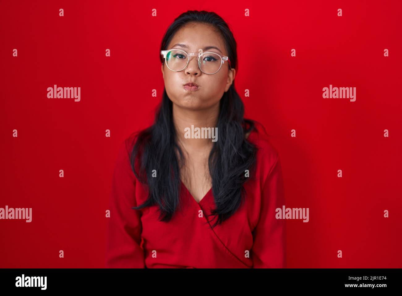 Asian young woman standing over red background puffing cheeks with ...