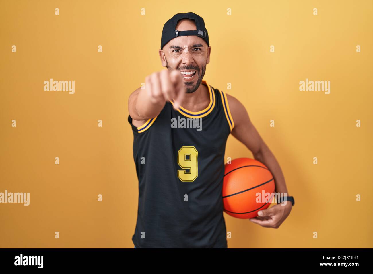 Middle age bald man holding basketball ball over yellow background ...