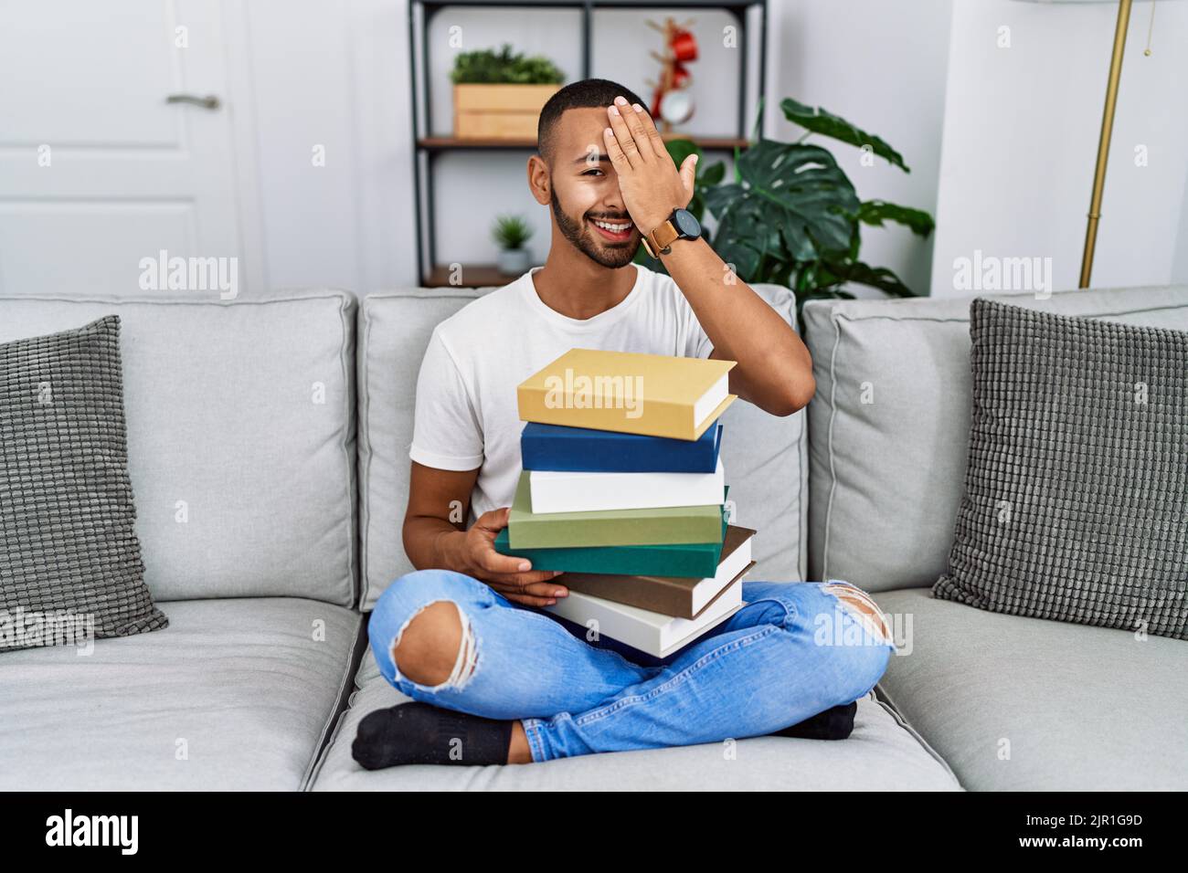 African american young man holding a pile of books sitting on the sofa ...