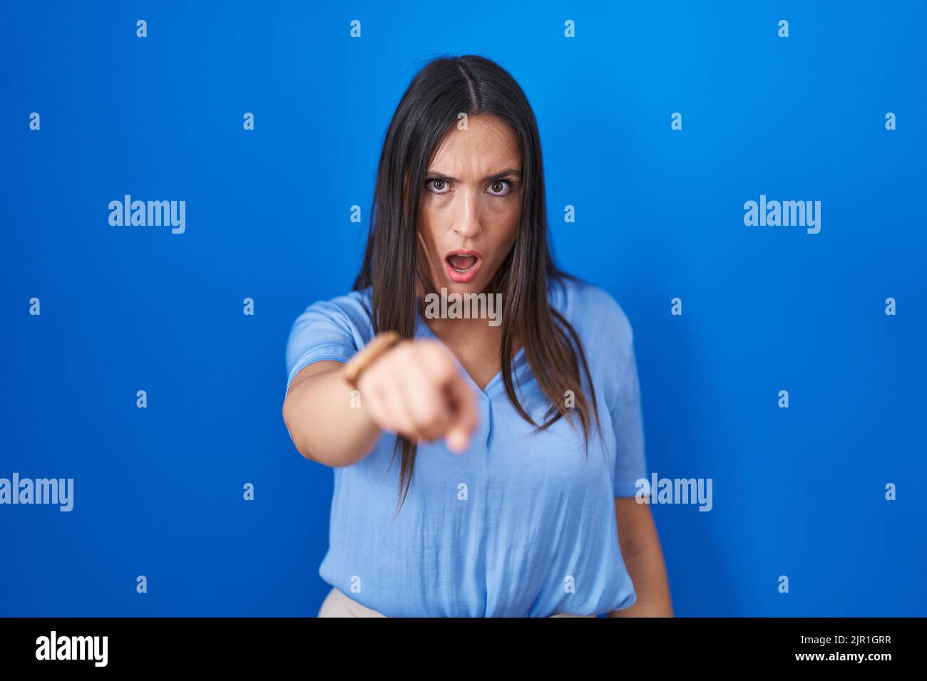 Young brunette woman standing over blue background pointing displeased ...