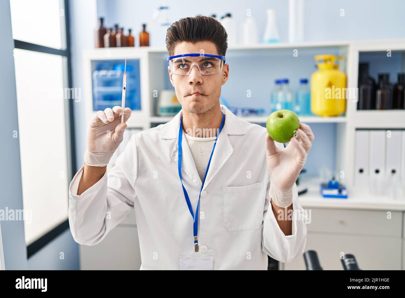 Young hispanic man working at scientist laboratory holding apple making ...