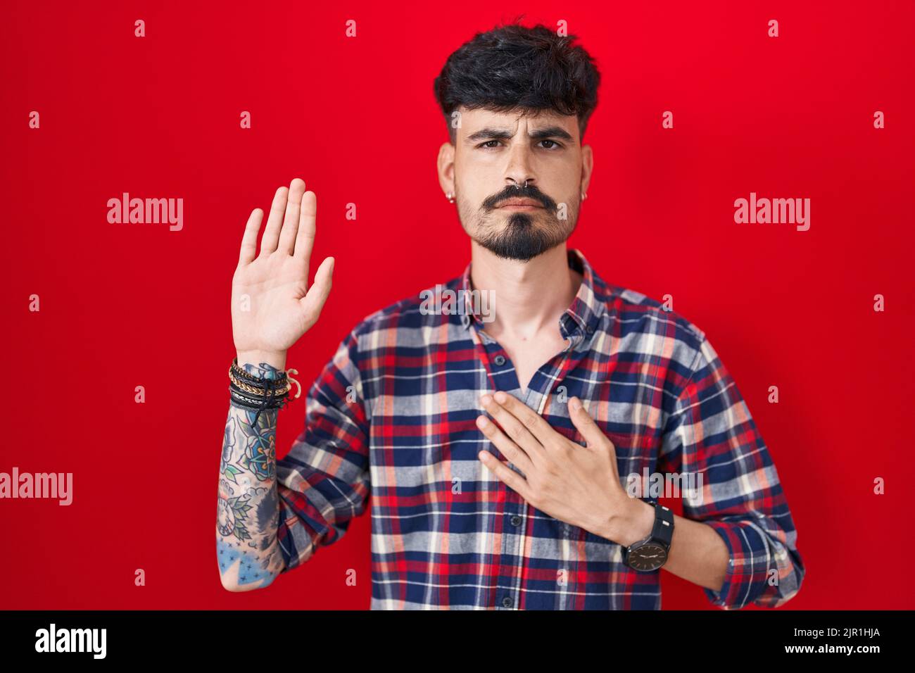 Young hispanic man with beard standing over red background swearing ...