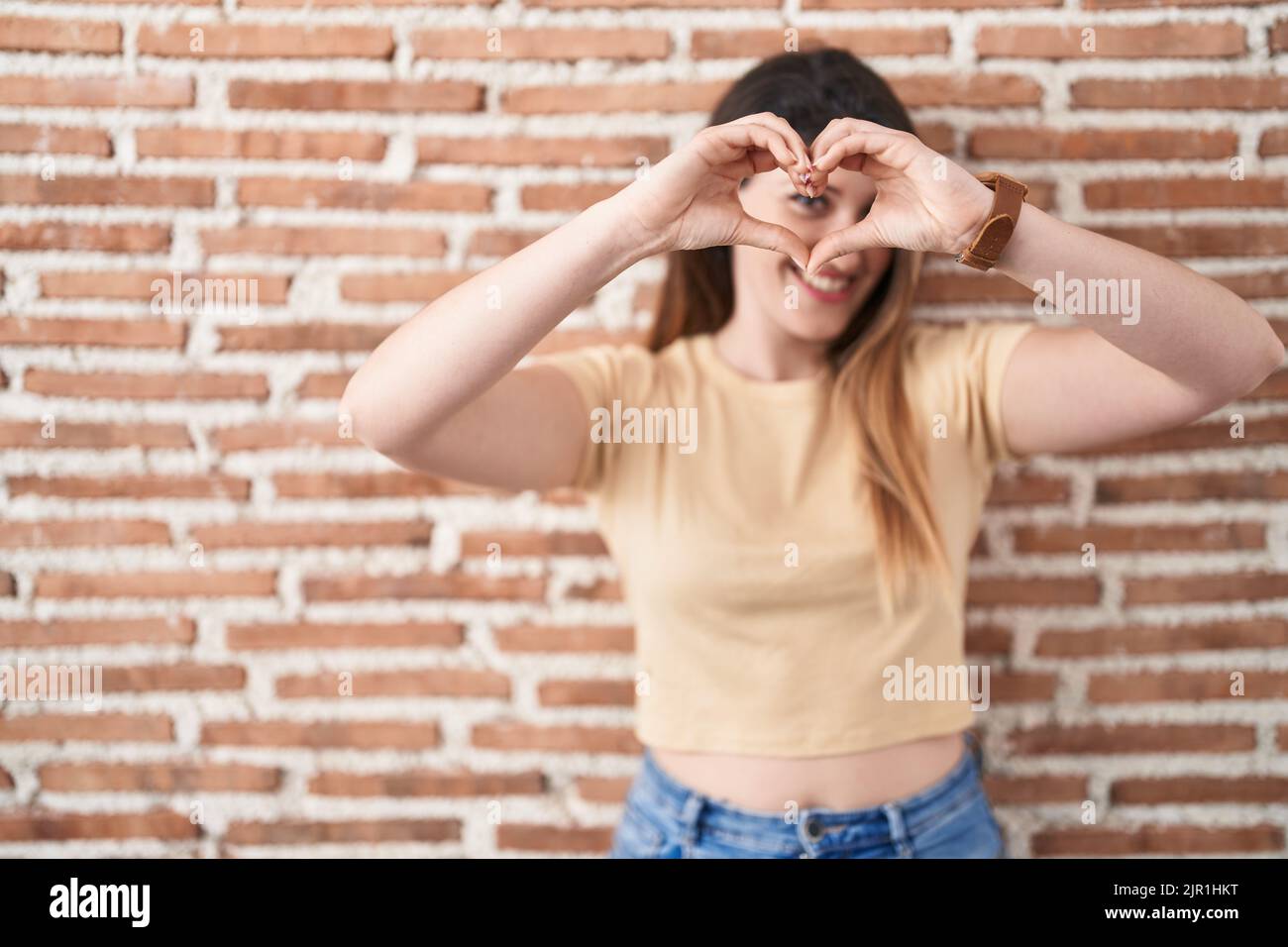Young brunette woman standing over bricks wall doing heart shape with ...