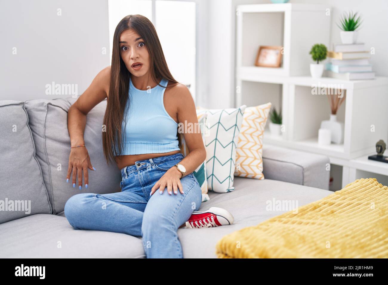 Young brunette woman sitting on the sofa at home in shock face, looking ...