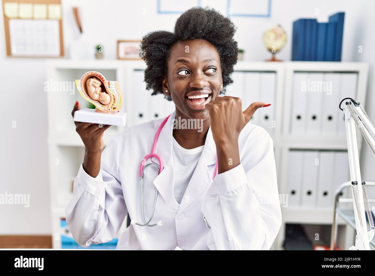 African doctor woman holding anatomical model of female uterus with fetus pointing thumb up to ...
