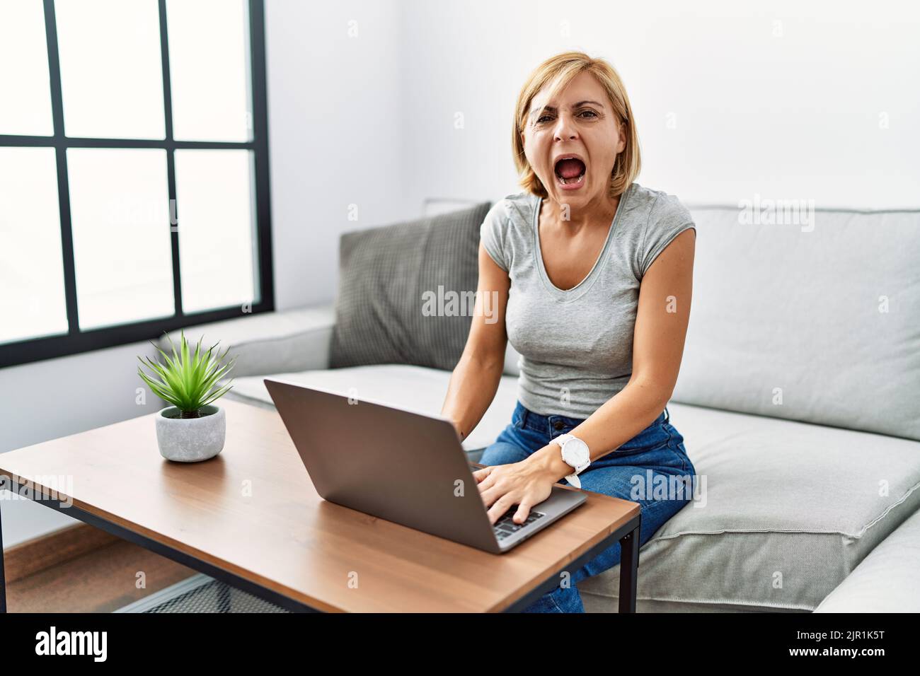 Middle age blonde woman using laptop at home angry and mad screaming ...