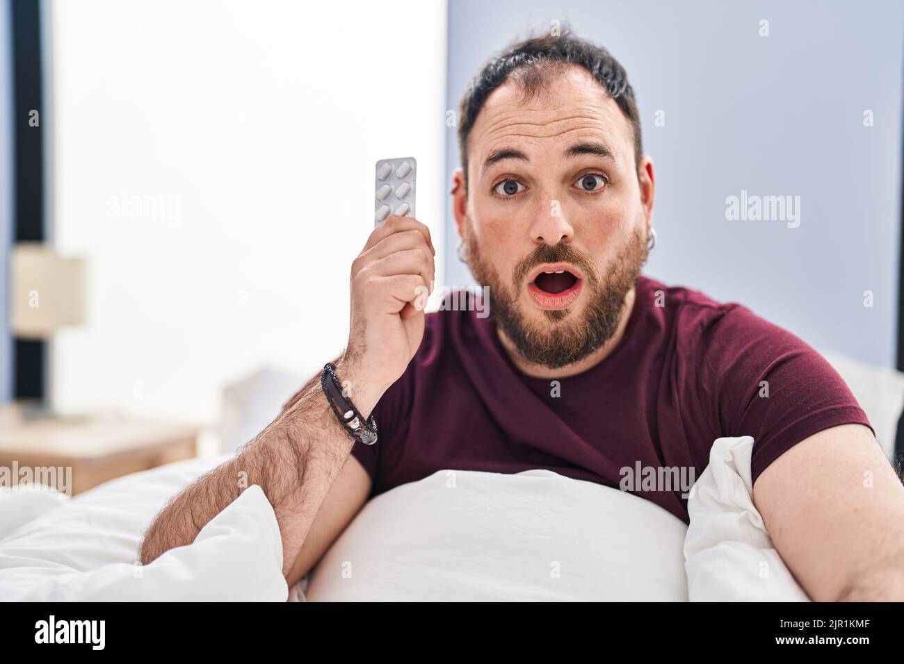 Plus size hispanic man with beard in the bed holding pills scared and ...