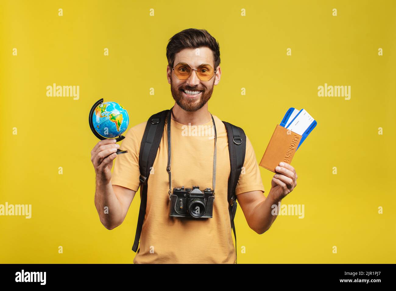 Happy man with passport, tickets, globe, camera and backpack posing over yellow studio ...