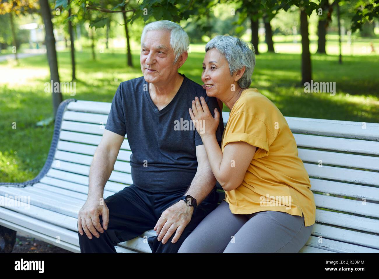 Pretty multi racial elderly 60s couple resting sit on bench in park after stroll, morning walk ...