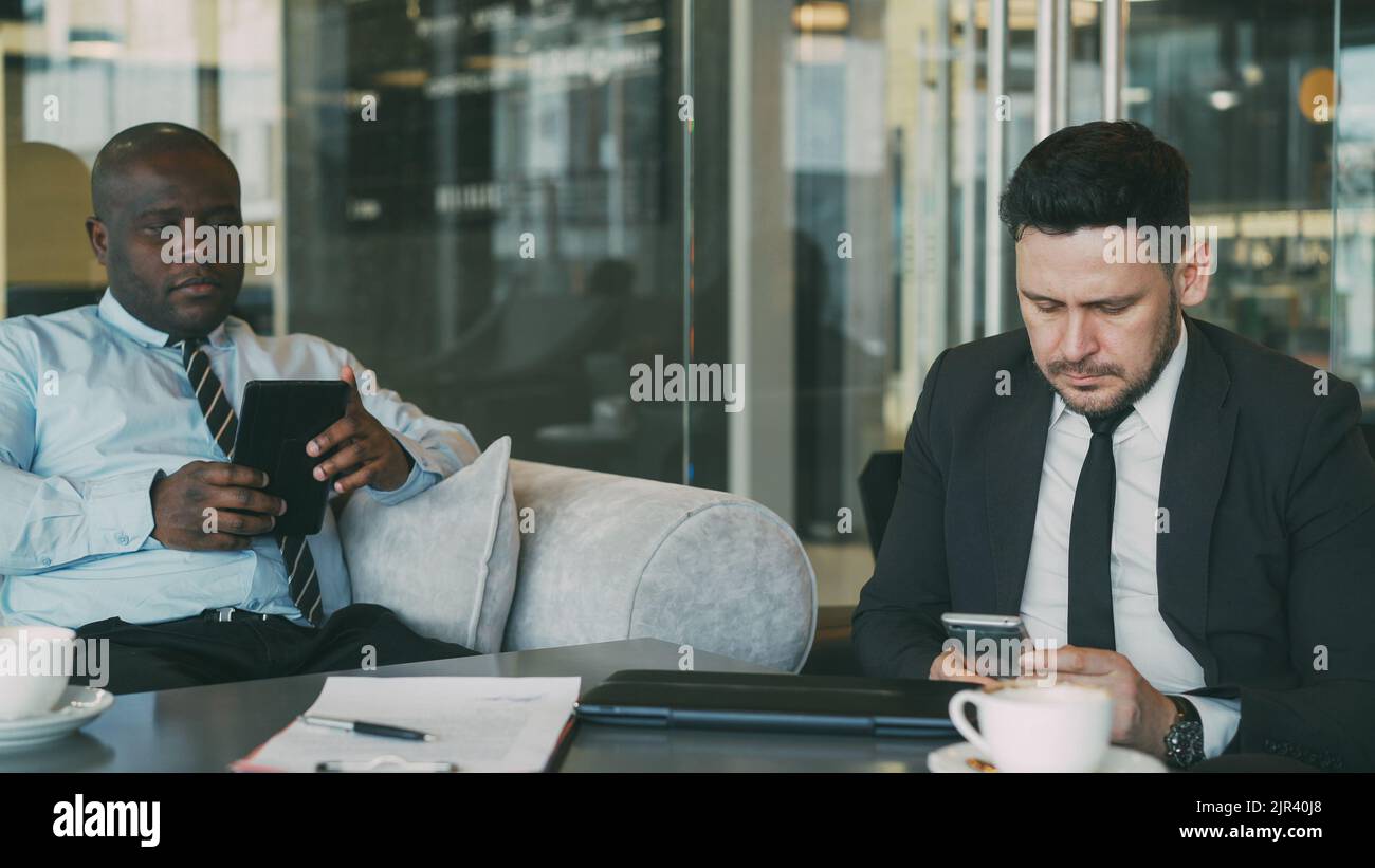 Two multi-ethnic businessmen in formal clothes sitting and using their ...