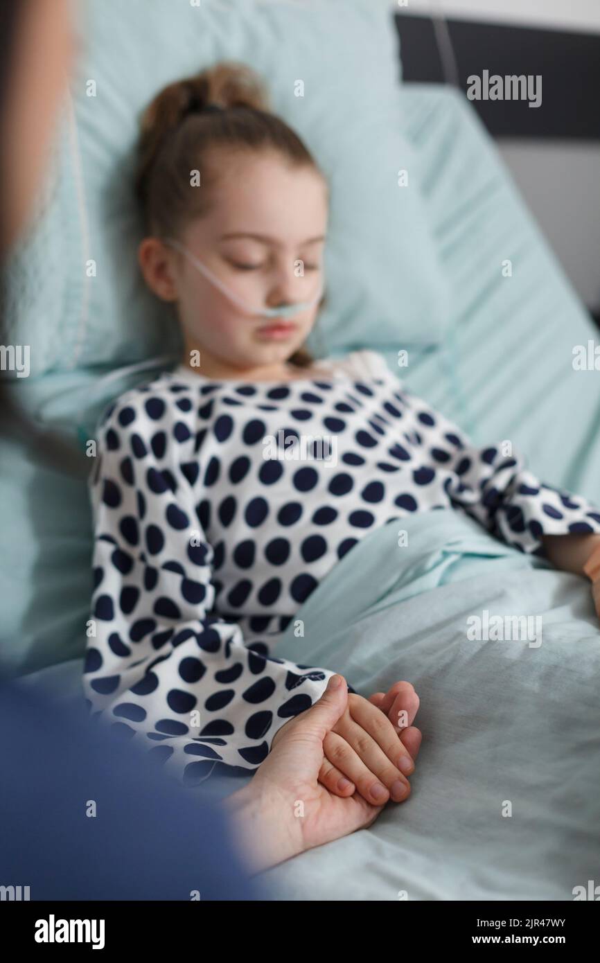 Hospitalized sick girl under treatment sleeping on patient bed while mother holding her hand ...