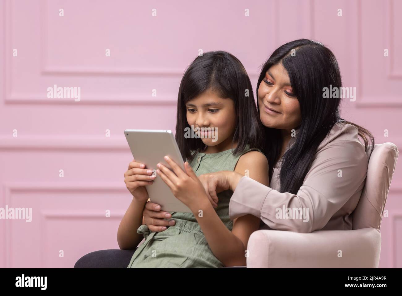 A Hispanic mother with her daughter using a tablet on a pink background ...