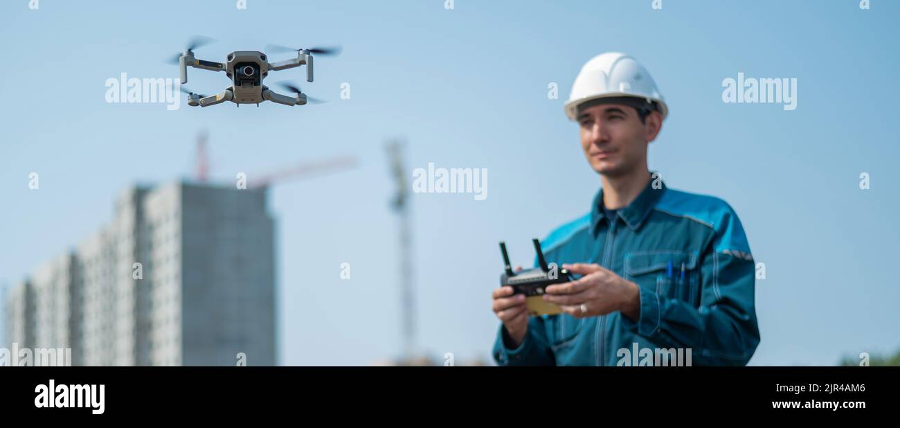 A man in a helmet and overalls controls a drone at a construction site ...