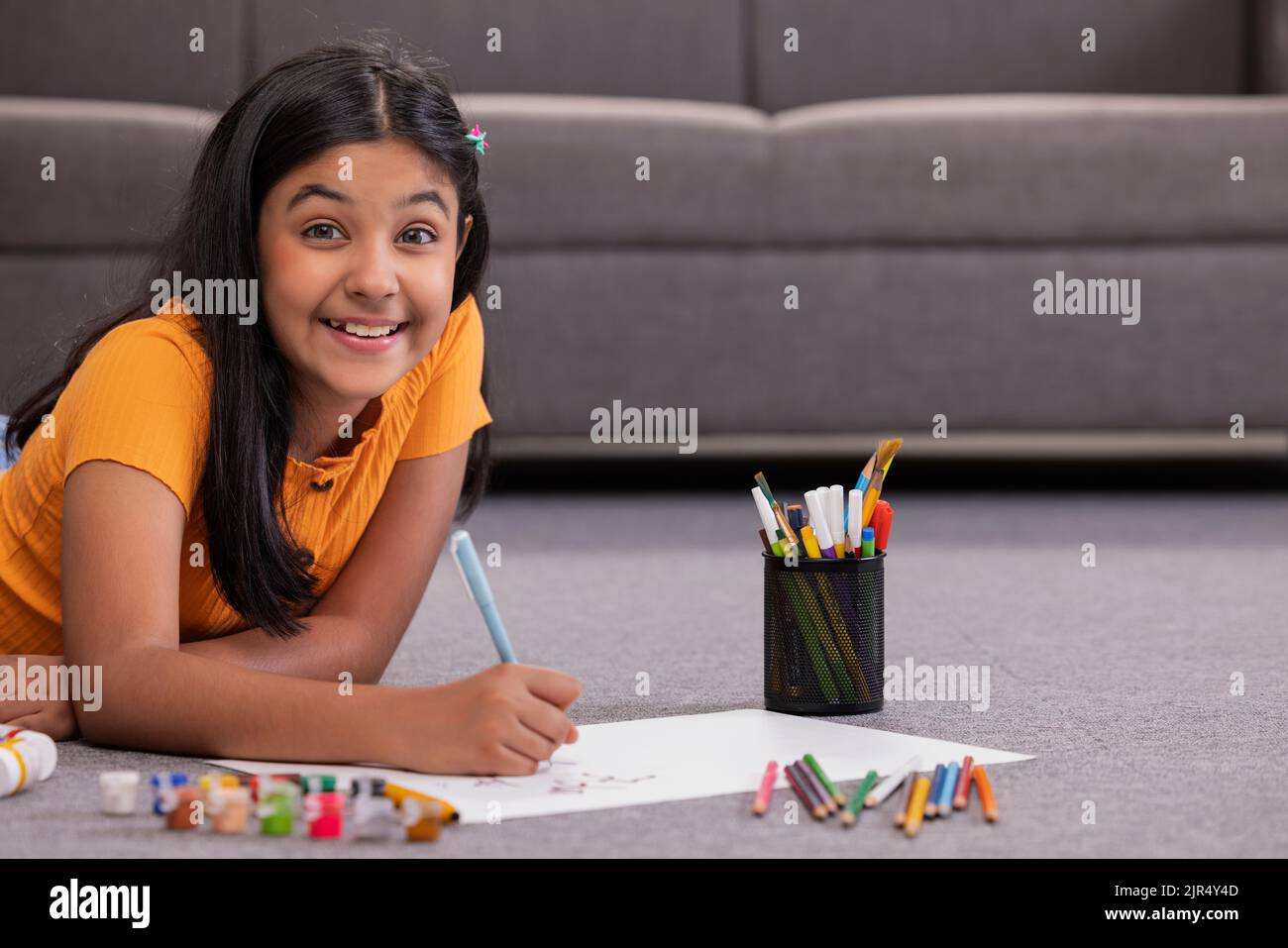 Portrait of a girl drawing on paper while lying down on floor in living ...