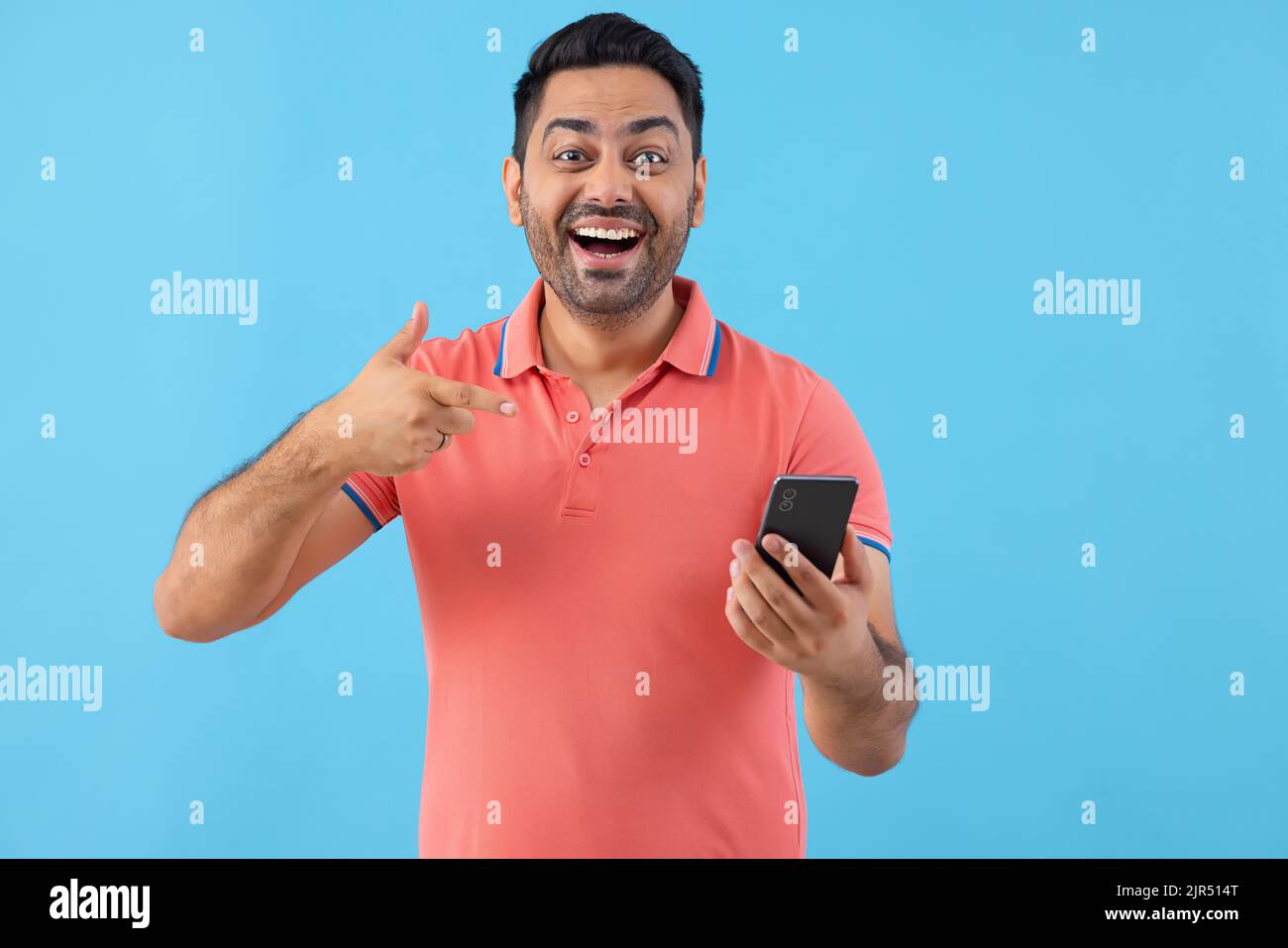 Smiling young man looking at camera and pointing at Smartphone holding ...