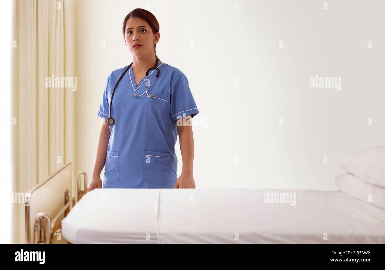Portrait of a young nurse standing beside patient bed in hospital ward ...