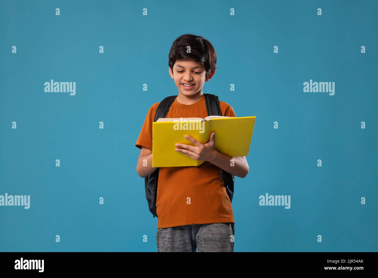 Boy reading book while standing with backpack against blue background ...