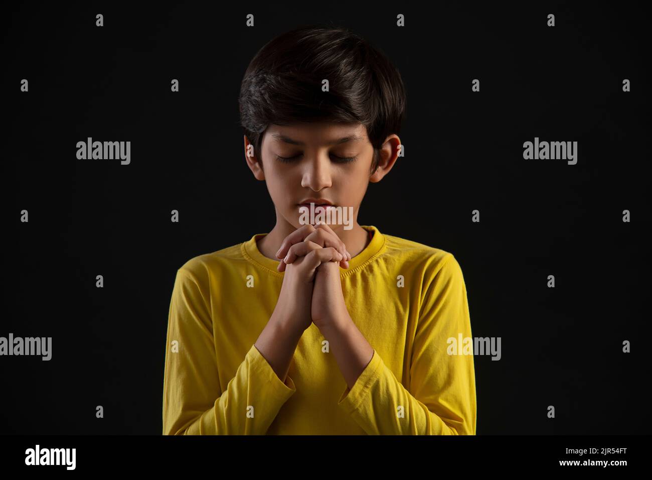 Boy with hands clasped praying while standing against dark background ...