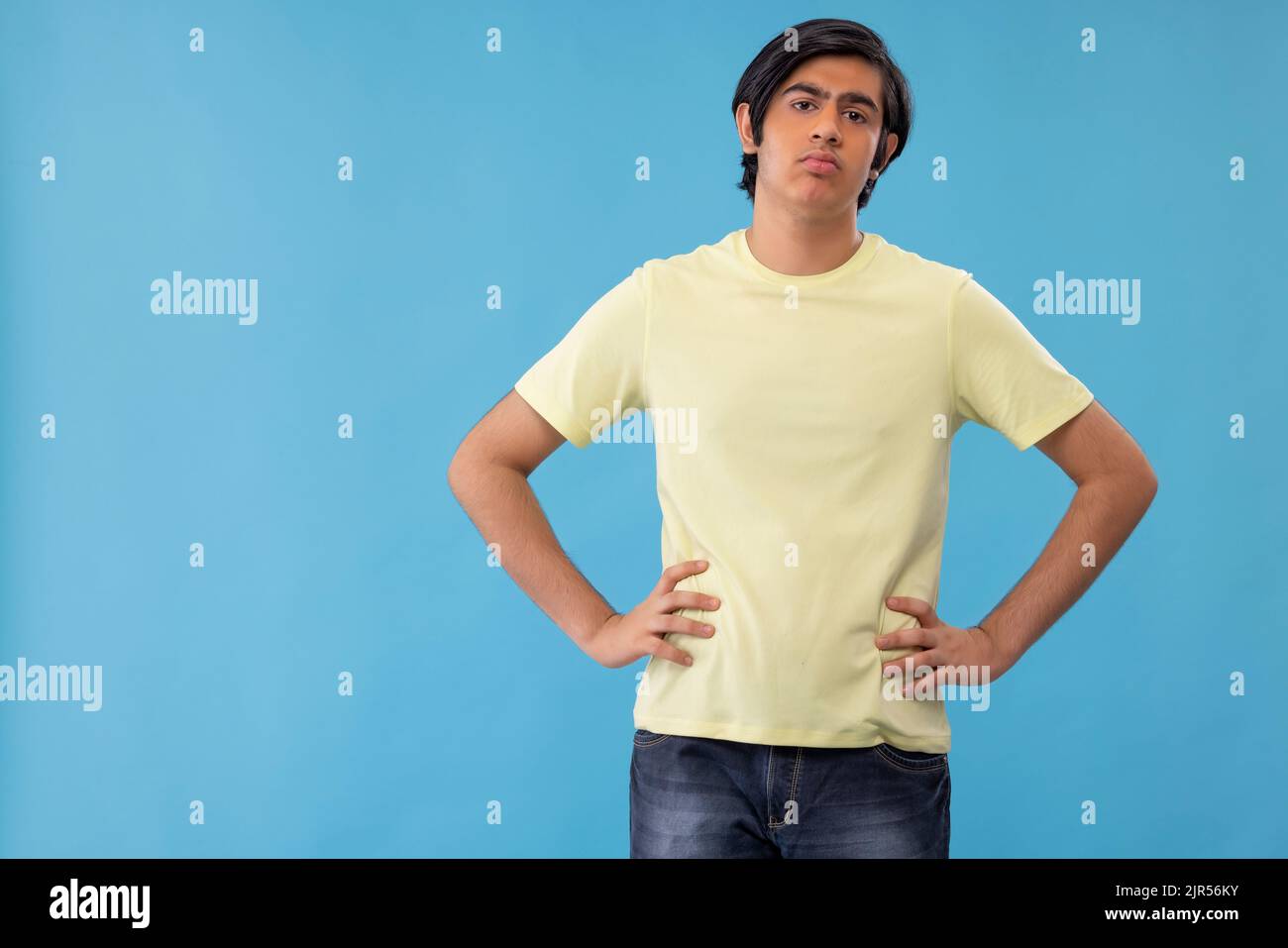 Portrait of a sad teenage boy with hands on waist standing against blue ...