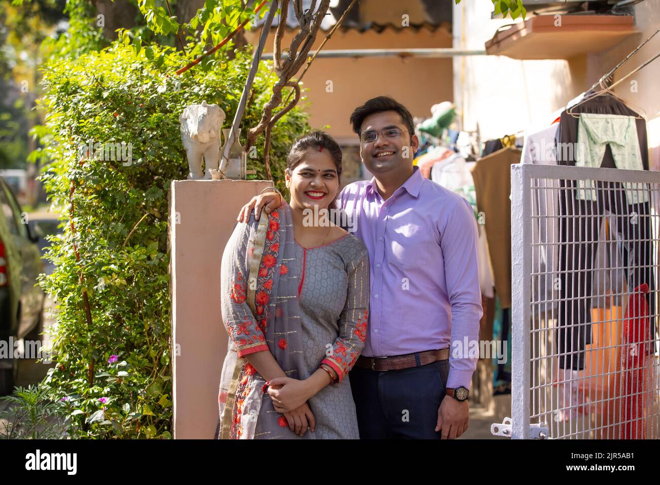 Portrait of young couple standing in front of gate with arm on shoulder ...