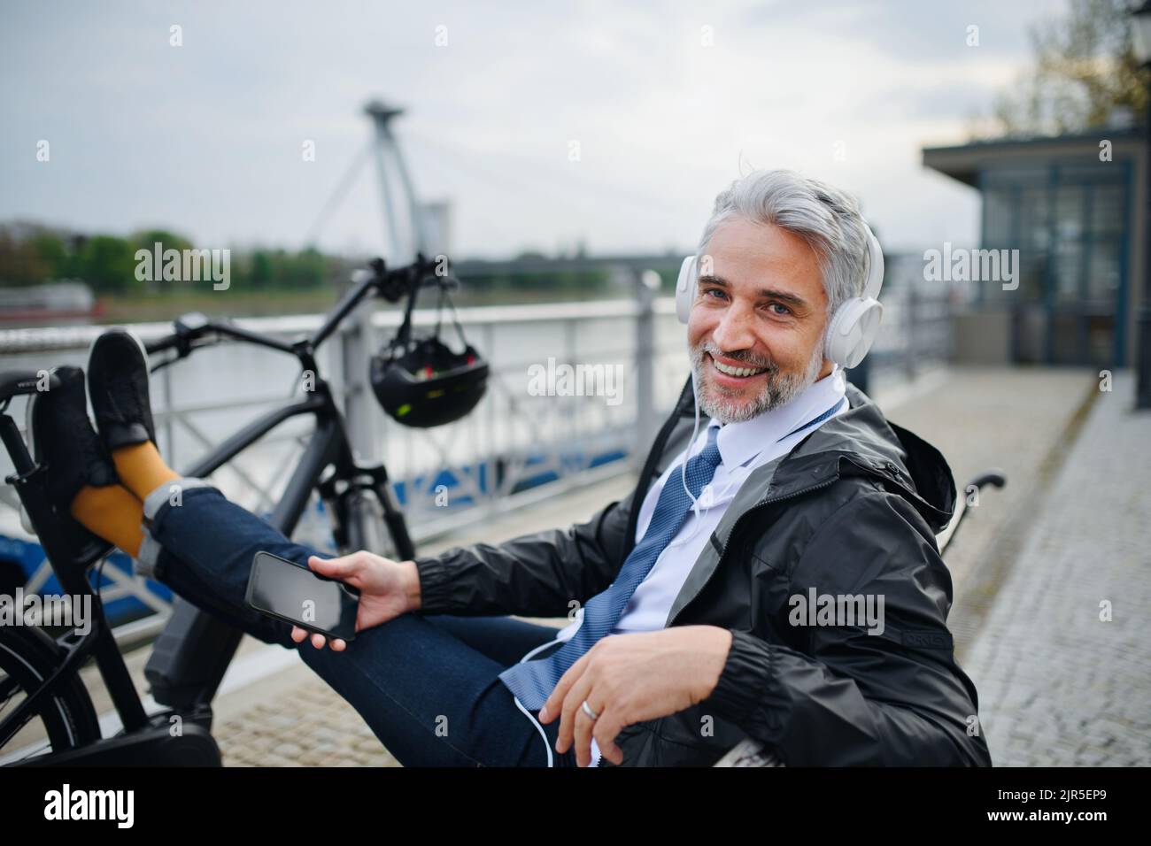Businessman with bike sitting on bench, listening to music with feet up ...